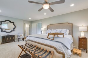 Um quarto com cama, ventoinha no teto, mesa de cabeceira, cômoda, cadeira e um pequeno banquinho.