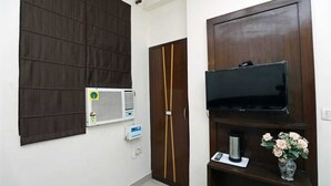 Quarto com uma grande janela, ar condicionado, porta de madeira, televisão sobre um móvel de madeira e uma pequena mesa com um vaso de flores.