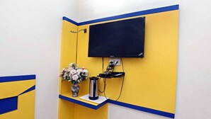 Quarto com esquema de cores amarelo e azul, uma TV de tela plana na parede, um vaso com flores e um objeto preto sobre uma superfície branca.