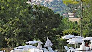 Terraço com cadeiras de descanso e guarda-sóis, com vista para uma vila situada no topo de uma colina.