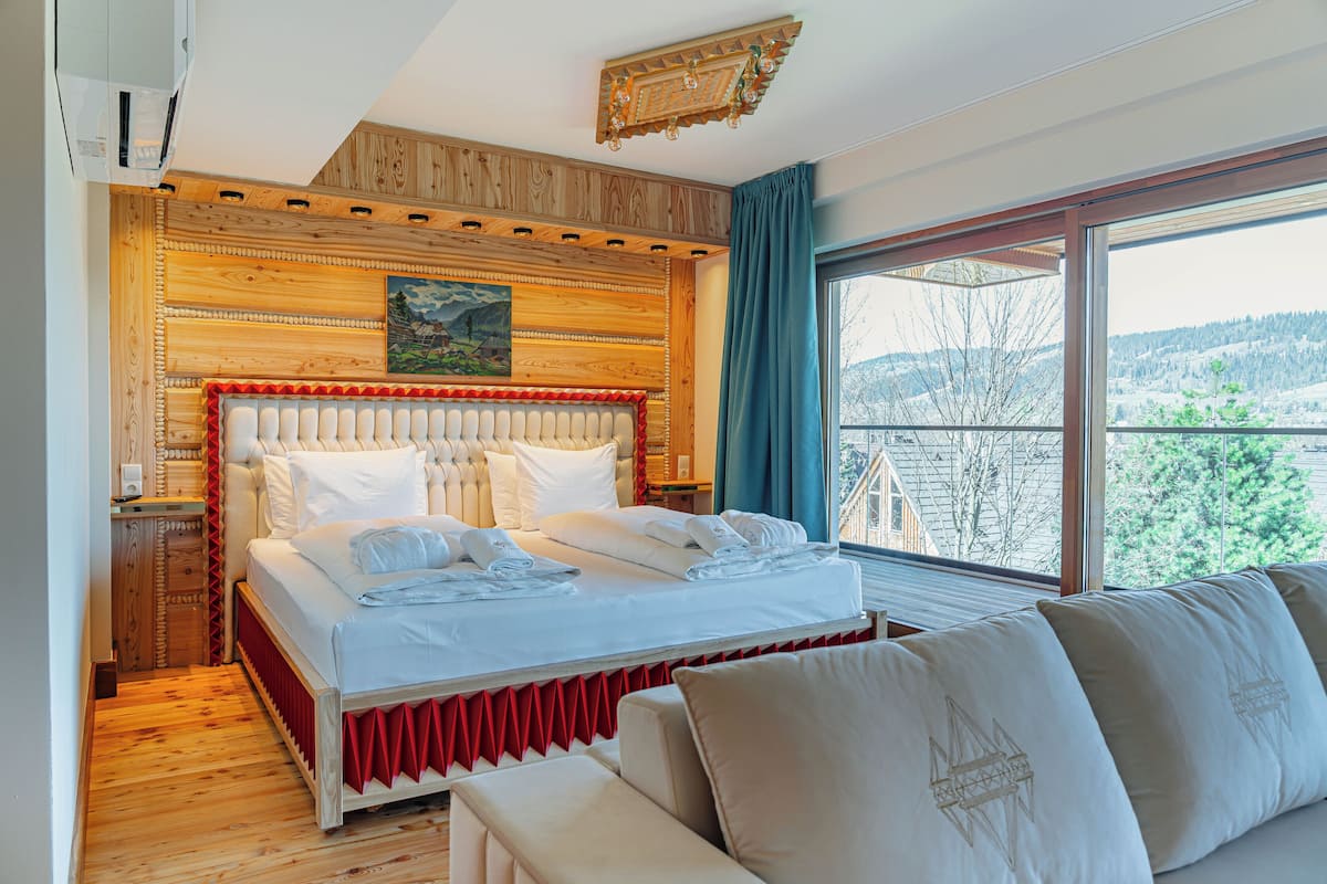 Quarto com cabeceira de madeira, uma janela grande com vista para árvores e cama com roupa de cama branca.