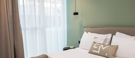 Cama bem arrumada com manta bege e toalhas, travesseiro com logotipo, mesa de cabeceira com abajur e cortinas leves.