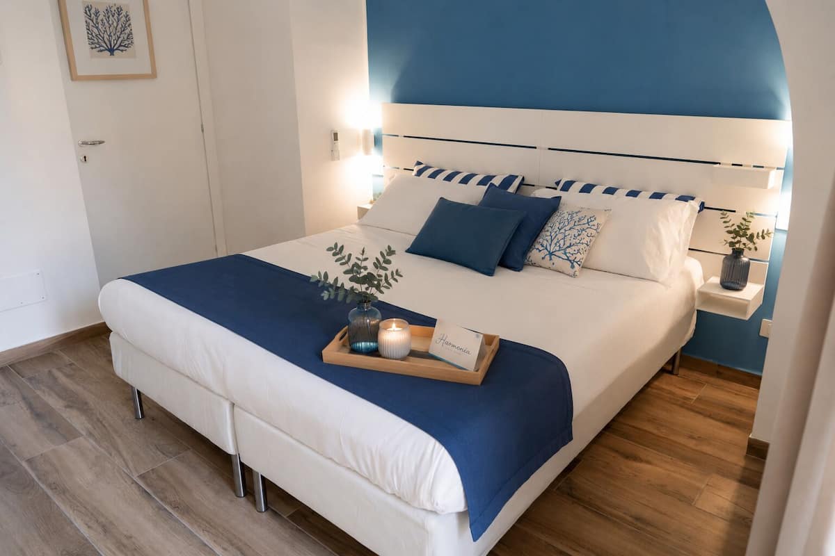 Uma cama bem arrumada com esquema de cores azul e branco, uma mesa de cabeceira com um vaso e uma vela, e um quadro na parede.