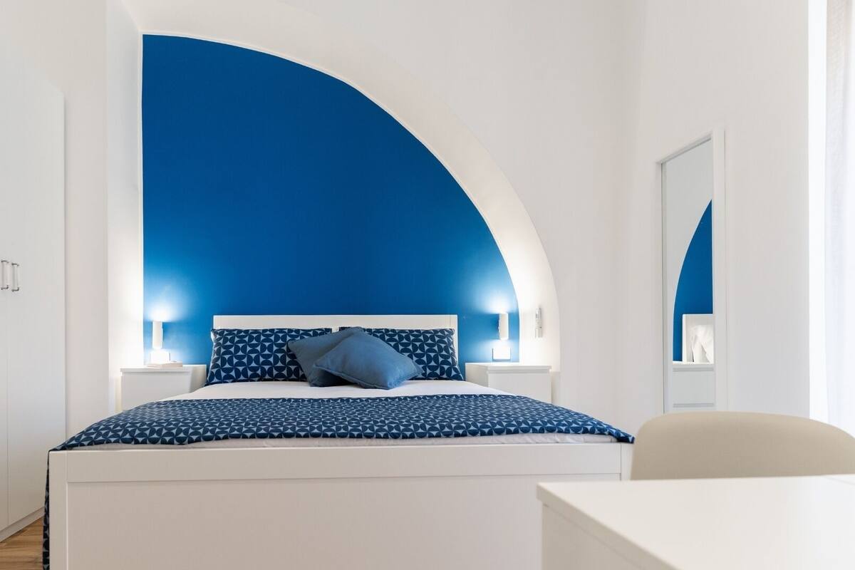 Quarto moderno com teto arredondado, cama com roupa de cama estampada, abajures e escrivaninha branca.