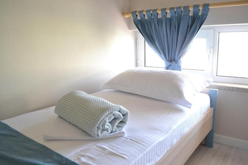 Uma cama de solteiro com colchão e almofadas brancos, um cobertor enrolado e uma janela com cortina azul.