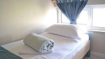 Uma cama de solteiro com colchão e almofadas brancos, um cobertor enrolado e uma janela com cortina azul.