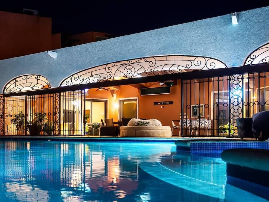 Piscina cercada, área de descanso com cadeiras e um sofá, e um edifício com entrada em arco marcante.