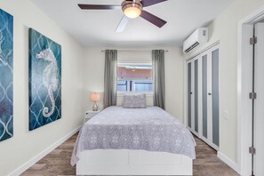 Quarto com cama, ventilador de teto, janela com cortinas, mesa de cabeceira com abajur e quadro de um cavalo-marinho na parede.