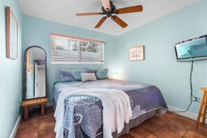 Quarto com cama, ventoinha no teto, televisão, janela com persianas e um banco de madeira.