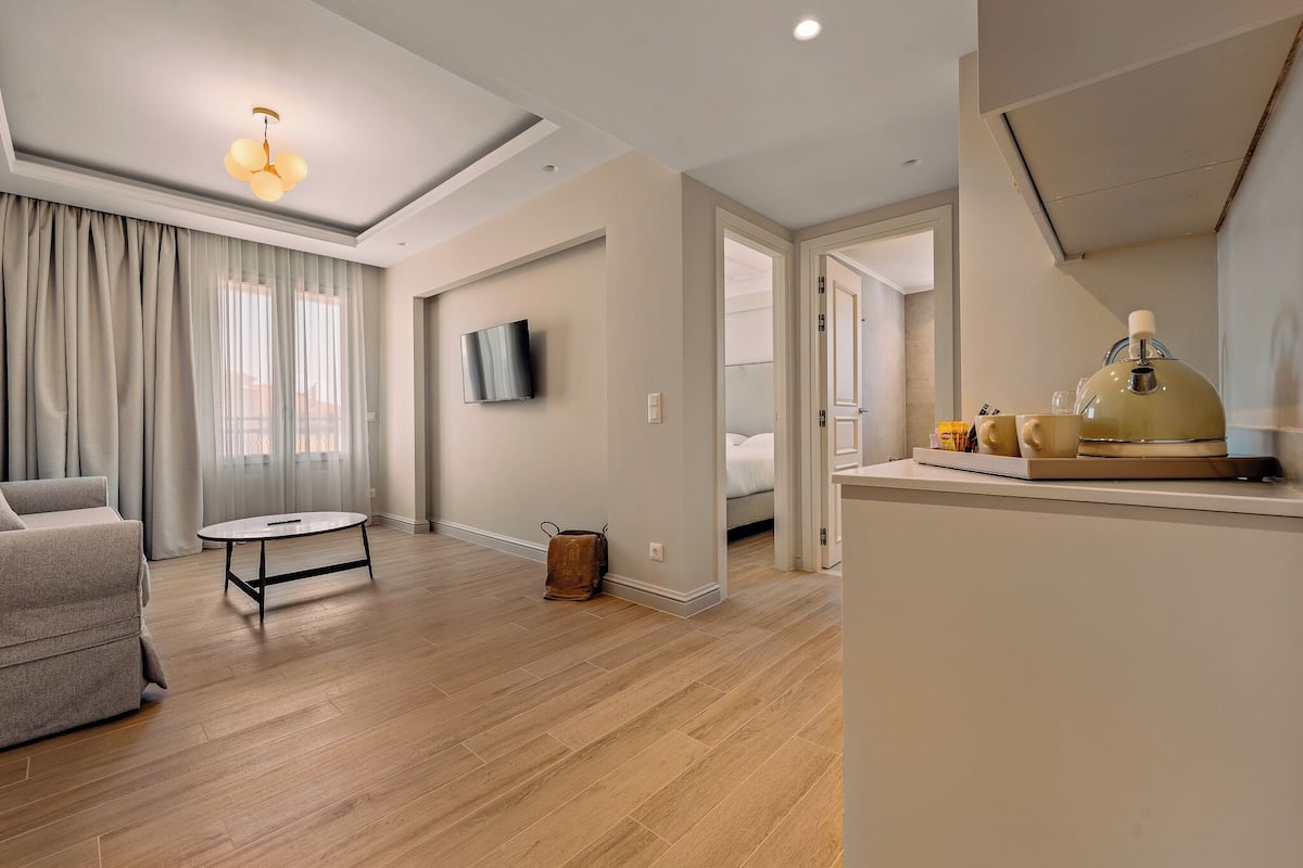 Quarto de hotel moderno com sofá, uma mesinha, televisão fixada na parede e uma área de kitchenette.