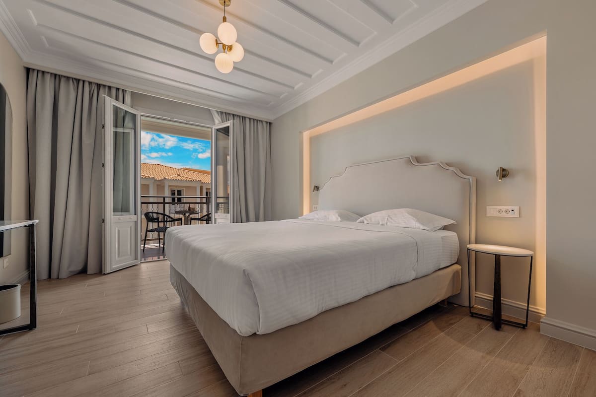 Quarto com cama, mesa de cabeceira, cadeira e vista através de uma porta aberta.