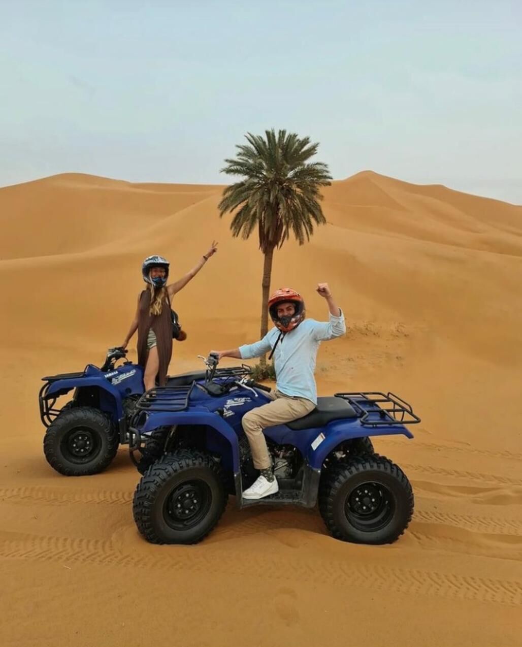 Duas pessoas em um veículo todo-terreno em um deserto, com dunas de areia e uma palmeira.