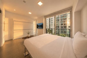 Quarto moderno com cama grande, televisão e varanda com vista para a cidade.