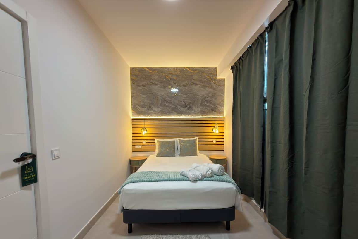 Quarto de hotel moderno com cama, cabeceira com iluminação embutida, mesinha de cabeceira pequena e vista exterior através de uma janela com cortinas.