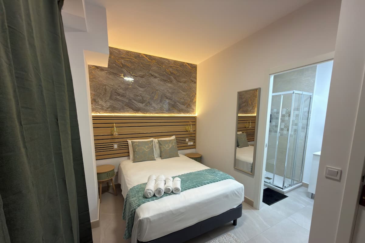 Quarto de hotel moderno com uma cama grande, mesa de cabeceira, espelho e chuveiro.
