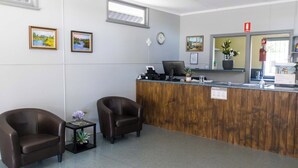 Uma área de recepção com uma mesa de madeira, duas poltronas de couro, uma pequena mesa lateral com uma planta, quadros nas paredes e um relógio.