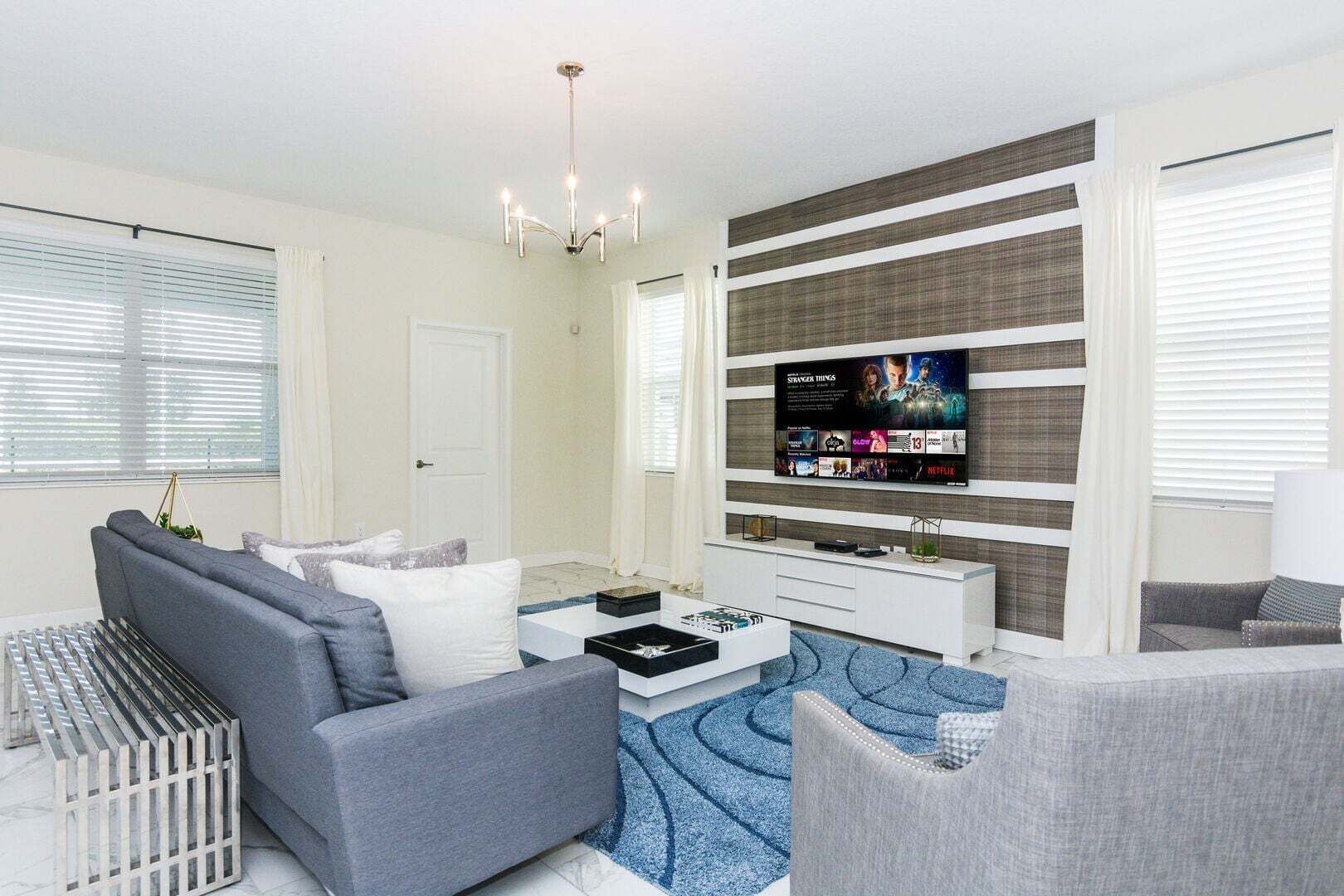 Uma sala de estar moderna com um sofá cinza, uma mesa de centro, uma TV fixada na parede e um tapete estampado.