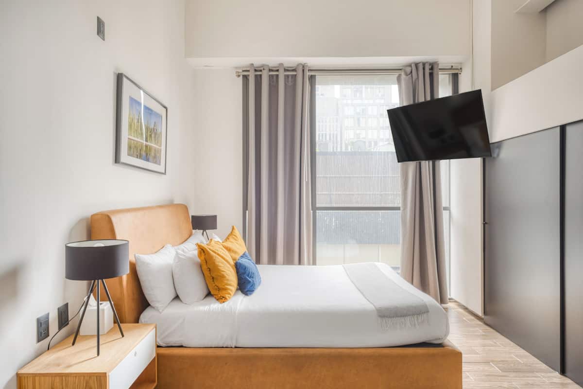 Quarto de hotel moderno com cama, mesa de cabeceira com abajur, televisão na parede e janela com cortinas.