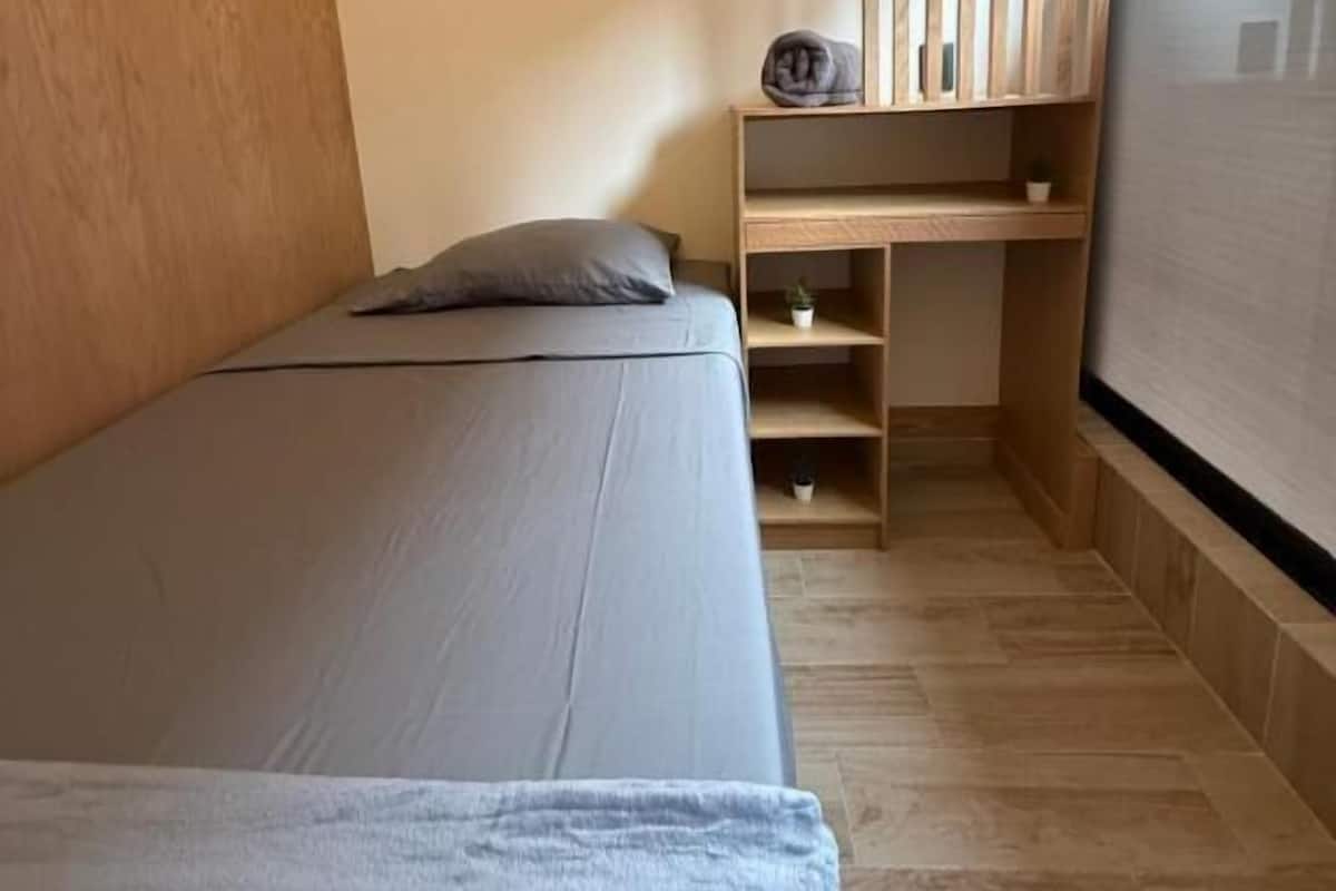 Uma cama de solteiro com um cobertor cinza, uma pequena mesa de cabeceira com abajur, uma estante de ripas de madeira e uma janela com persianas.