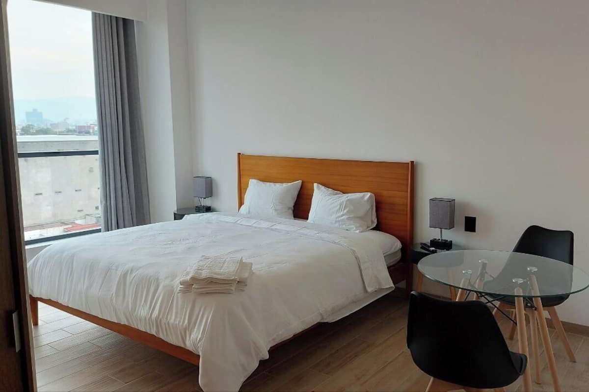 Quarto de hotel com cama, abajures, mesa de vidro com cadeiras e vista pela janela.