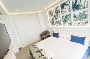 Quarto de hotel com cama, televisão, cadeira e janela com papel de parede tropical.