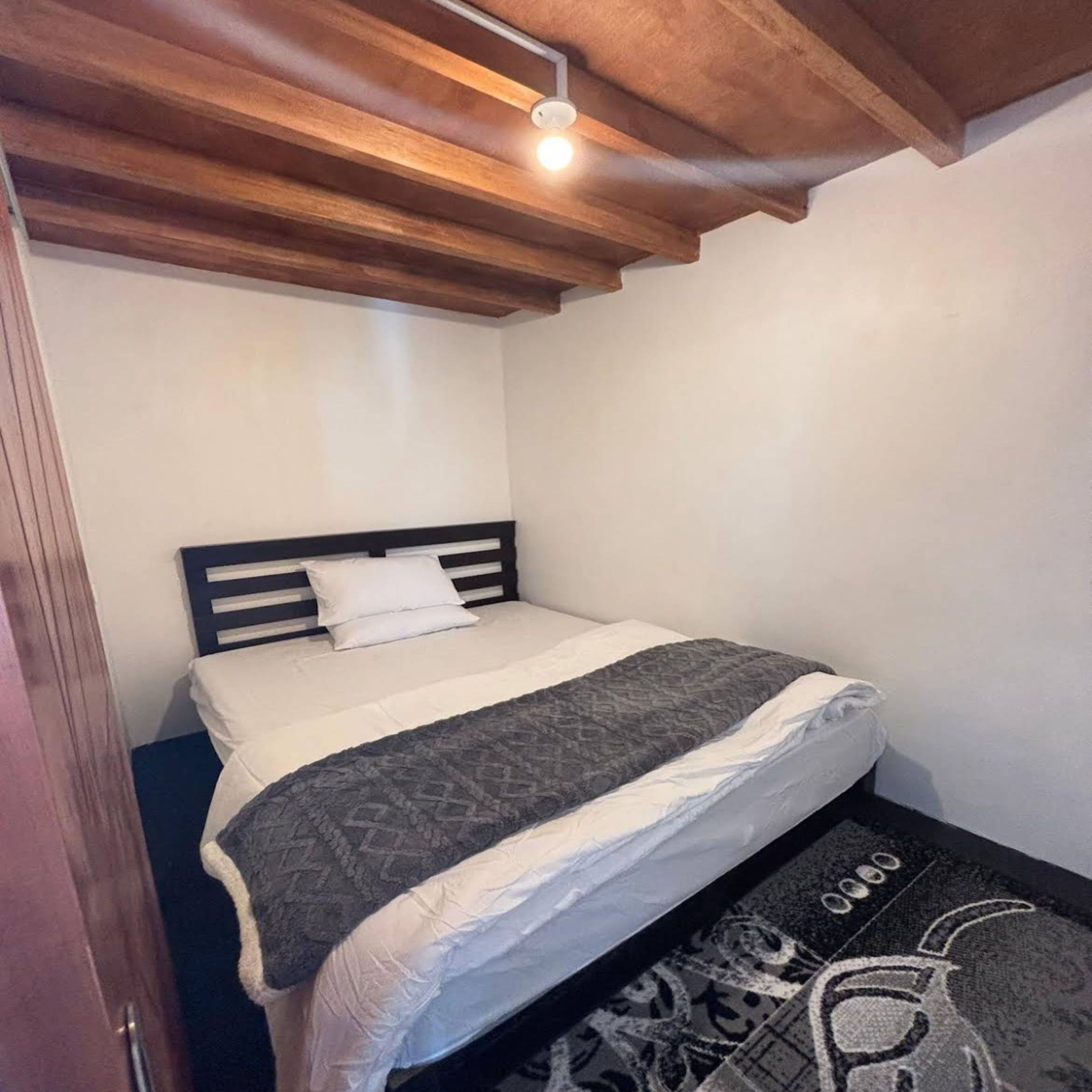 Quarto com teto de madeira, cama com roupa de cama branca e cinza, e tapete estampado.