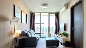 Quarto de hotel moderno com sofá, escrivaninha, luminária, televisão e uma janela grande com vista para a cidade.