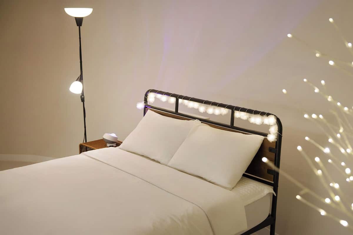 Uma cama com roupa de cama branca, uma mesa de cabeceira com abajur e uma guirlanda de luzes na parede.