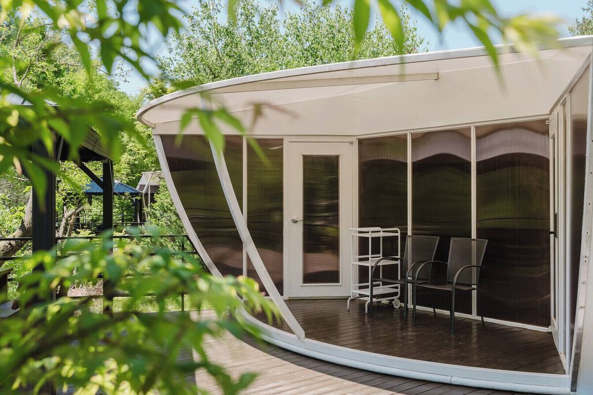 Um espaço moderno, com paredes de vidro curvo e um deck de madeira, uma cadeira de metal branca e uma mesa pequena.
