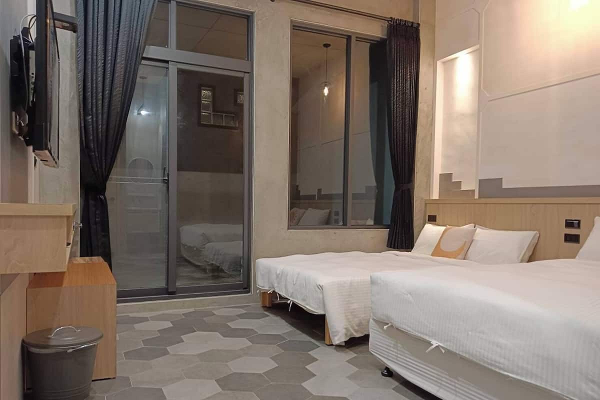 Quarto de hotel com duas camas, uma secretária de madeira pequena, um cesto de lixo e uma porta de vidro deslizante que dá para uma varanda.