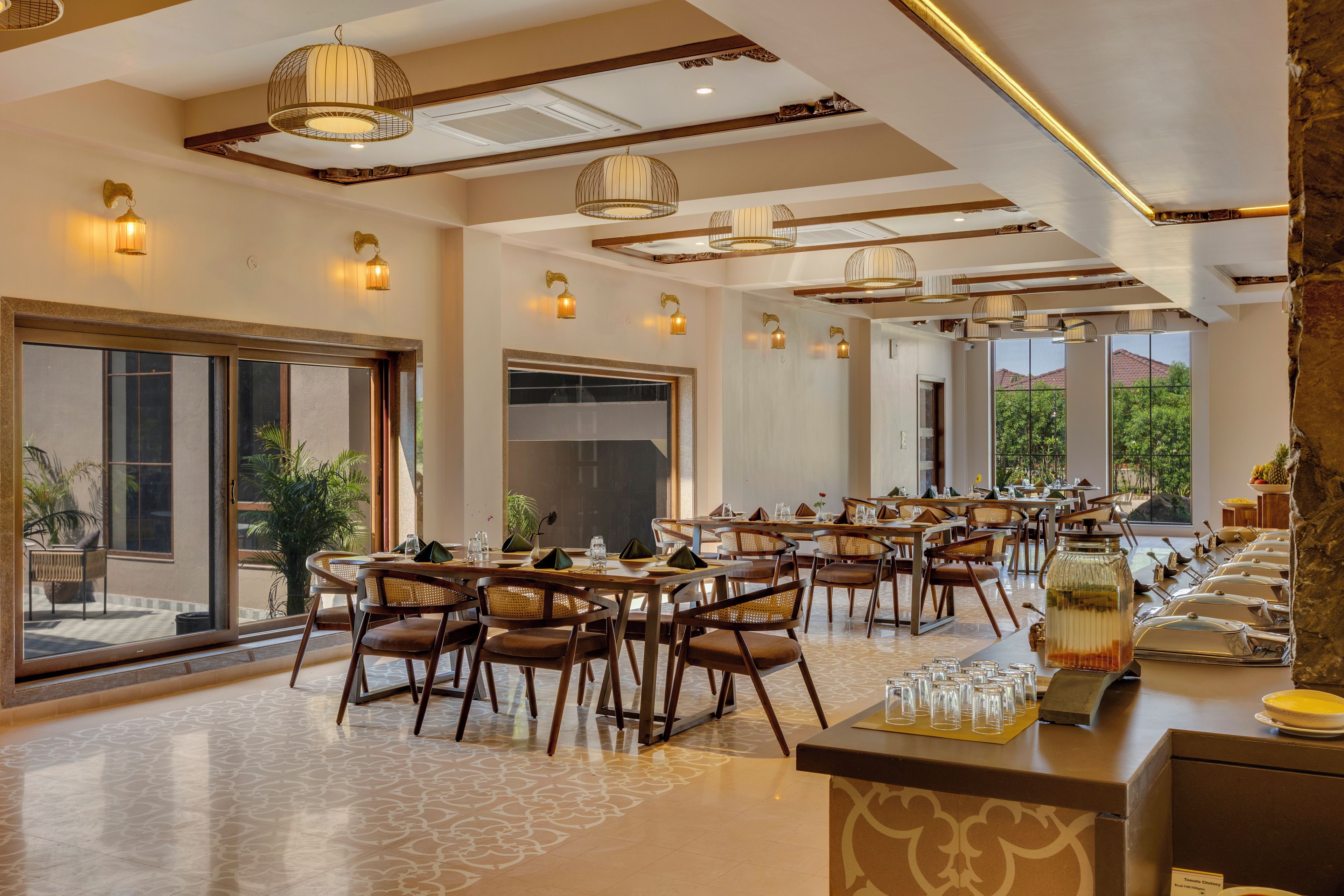 Uma ampla área de jantar com várias mesas e cadeiras, grandes janelas com vista para o exterior, e um buffet com diversos alimentos.