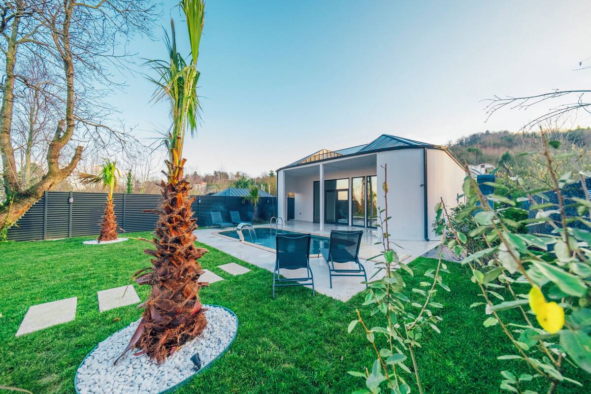 Uma casa moderna com piscina, cadeiras de patio e um gramado bem cuidado.