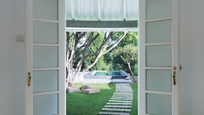 Visão através de uma porta branca com painéis de vidro fosco, que dá para um caminho pavimentado, uma pequena área gramada e uma piscina.
