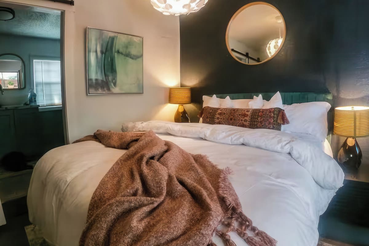 Quarto com uma cama grande, um espelho redondo, uma obra de arte na parede e um lustre de teto diferenciado.