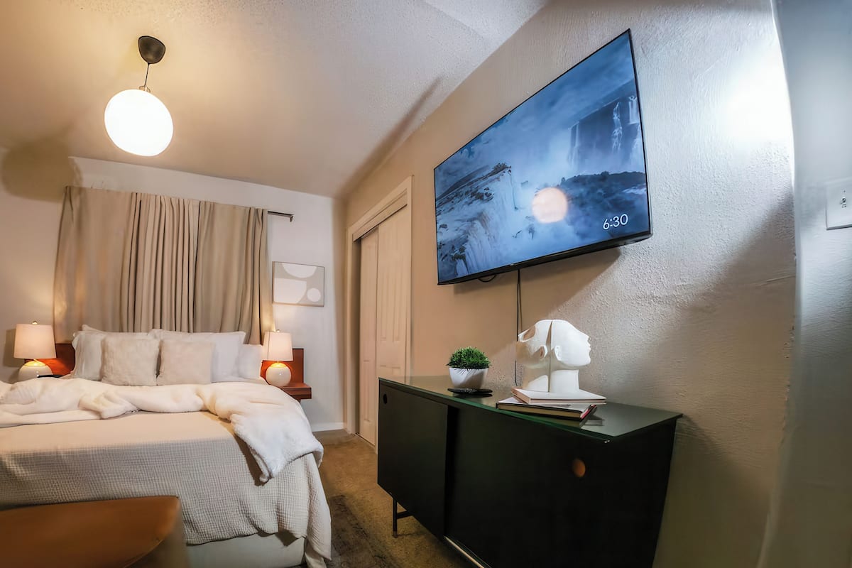 Quarto de hotel com cama, televisão na parede, mesa de cabeceira com abajur, cômoda e planta em vaso.