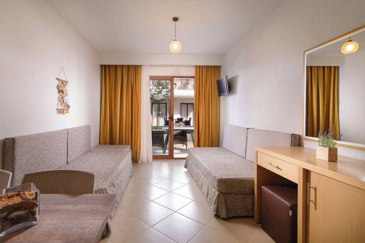 Quarto de hotel com duas camas, uma escrivaninha de madeira, um espelho, uma televisão e uma varanda com mesa e cadeiras.