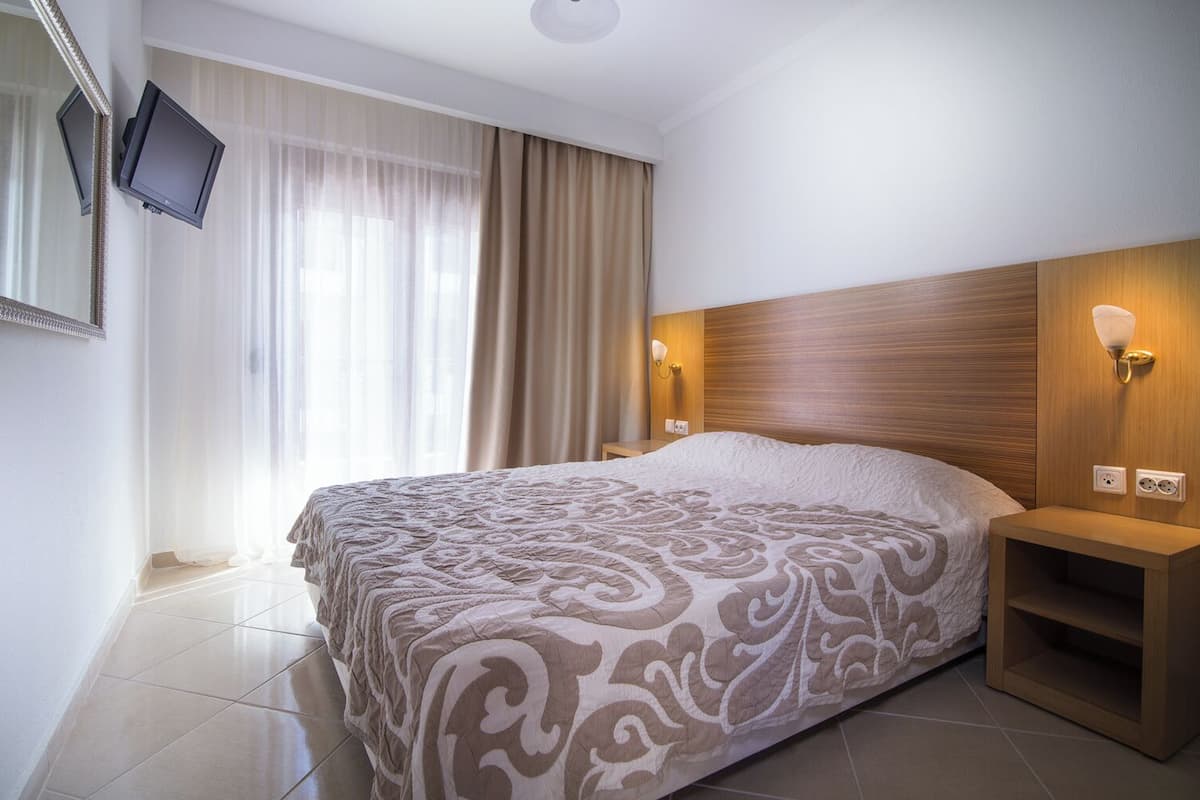 Quarto de hotel com uma cama grande, televisão fixada na parede, mesa de cabeceira e cabeceira de madeira.