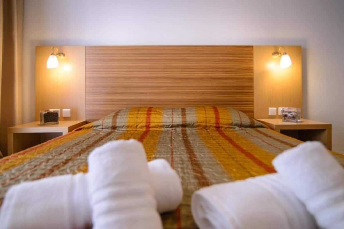 Quarto de hotel com uma cama, duas mesas de cabeceira e duas luminárias fixadas na parede.