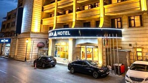 Um hotel com vários andares, entrada destacada, área de estacionamento com carros e céu claro.
