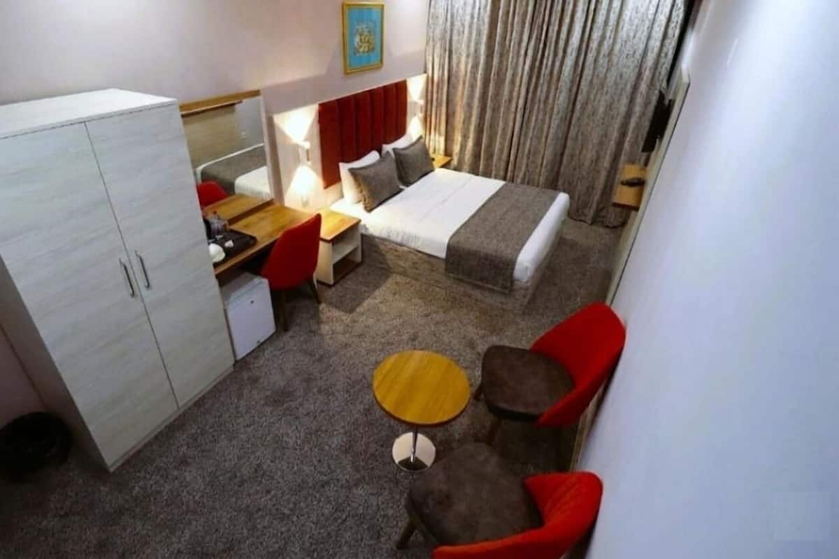 Quarto de hotel pequeno com uma cama, uma escrivaninha, um guarda-roupa e duas cadeiras vermelhas.