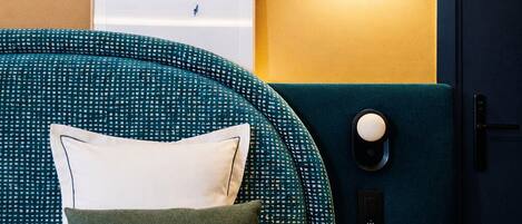 Quarto de hotel bem arrumado com cama, almofadas, mesa de cabeceira, abajur, porta e luminária na parede.