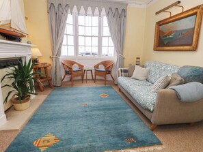 Uma sala de estar com um sofá, duas cadeiras de vime, um tapete azul, uma lareira e um quadro na parede.