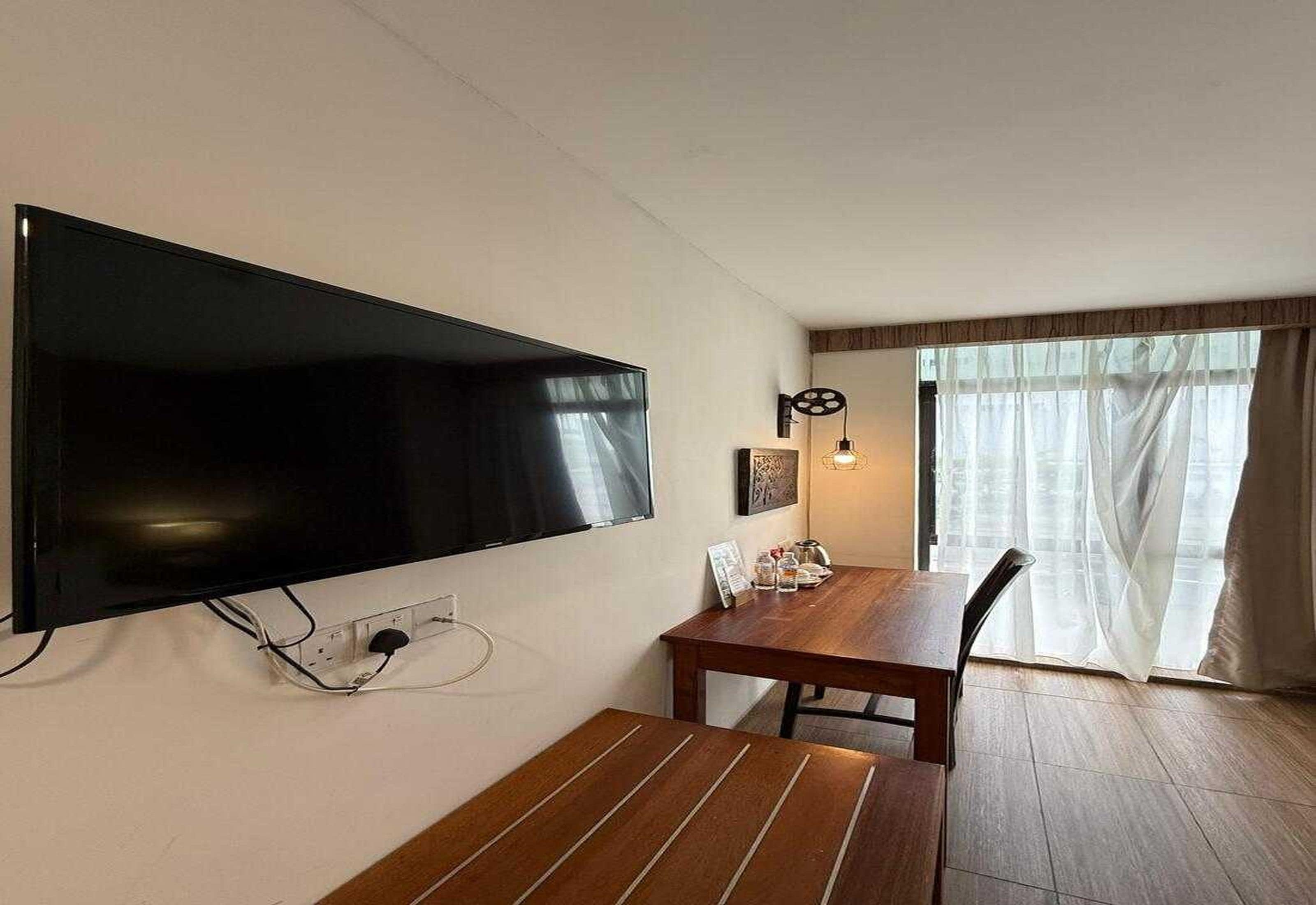 Quarto com televisão de tela plana na parede, mesa de jantar de madeira com cadeiras e janela com cortinas leves.