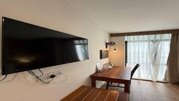 Quarto com televisão de tela plana na parede, mesa de jantar de madeira com cadeiras e janela com cortinas leves.