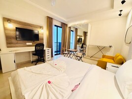 Quarto de hotel com cama, escrivaninha, cadeira, televisão e varanda com cortinas.