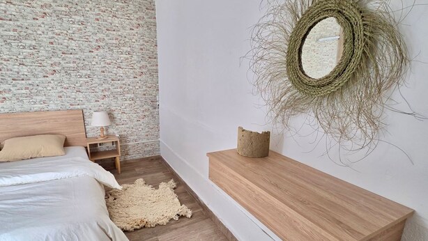 Quarto com cama, mesa de cabeceira, espelho fixado na parede, prateleira e um pequeno banquinho.