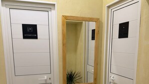 Corredor com duas portas brancas, cada uma identificada com a placa 'bathroom', um espelho e um vaso de planta.