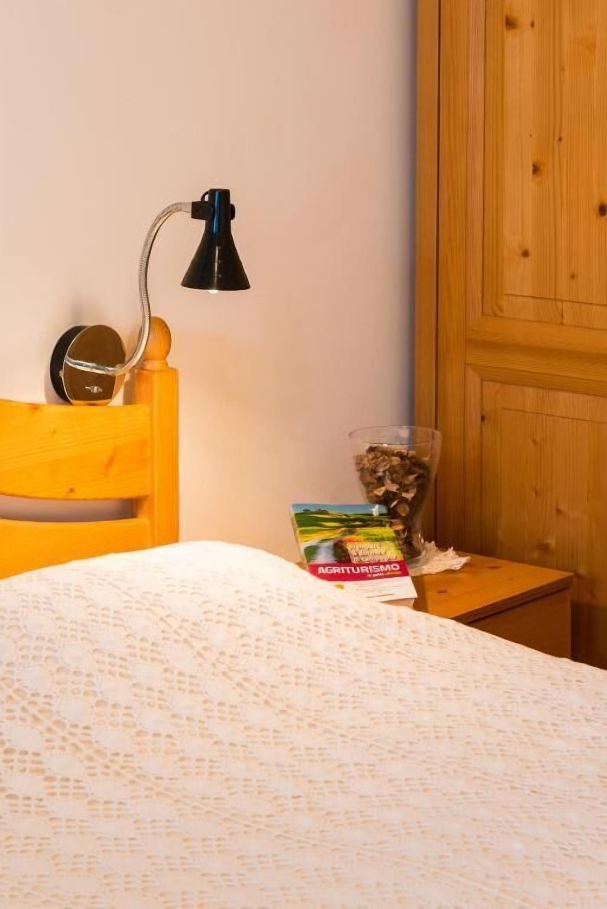 Quarto com cabeceira de madeira, uma escrivaninha com luminária, um livro e um recipiente de vidro.