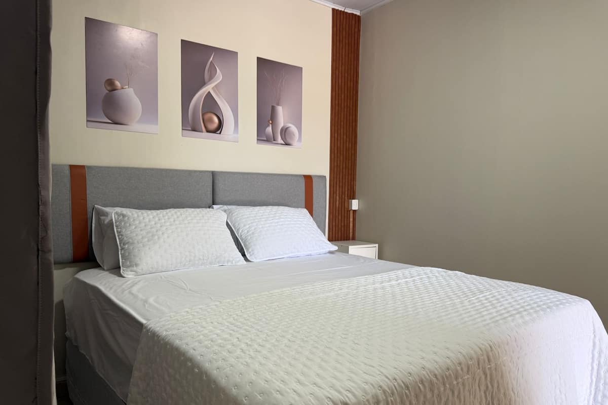 Uma cama bem arrumada com um edredom branco texturizado, cabeceira cinza e três obras de arte penduradas na parede retratando vasos e plantas abstratas.