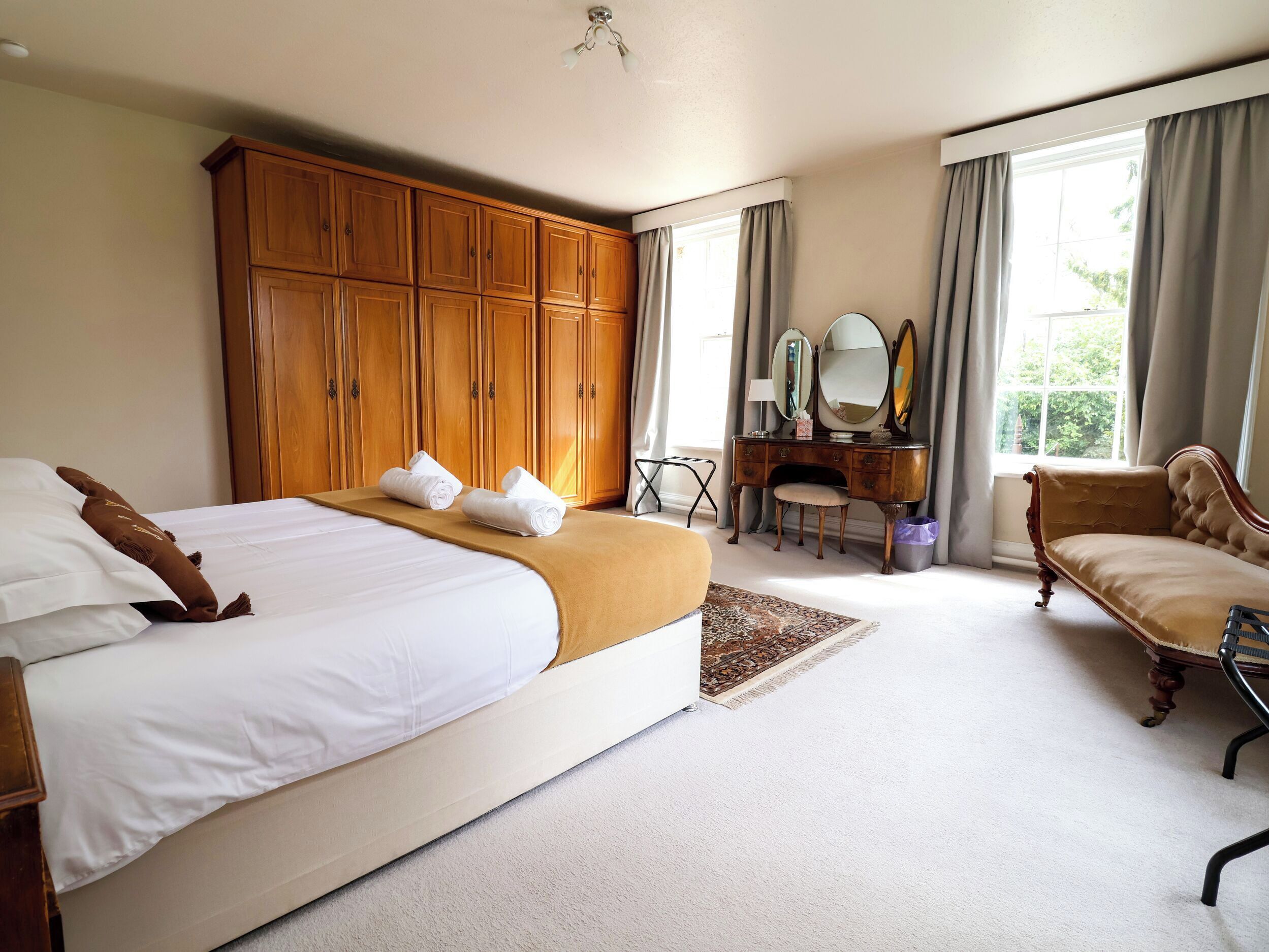 Quarto de hotel com cama grande, guarda-roupa de madeira, secretária com espelho e janela com cortinas.